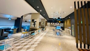 Um lobby de hotel moderno com piso de padrão geométrico, uma área de estar central com mesas e cadeiras, e um bar com banquetas.