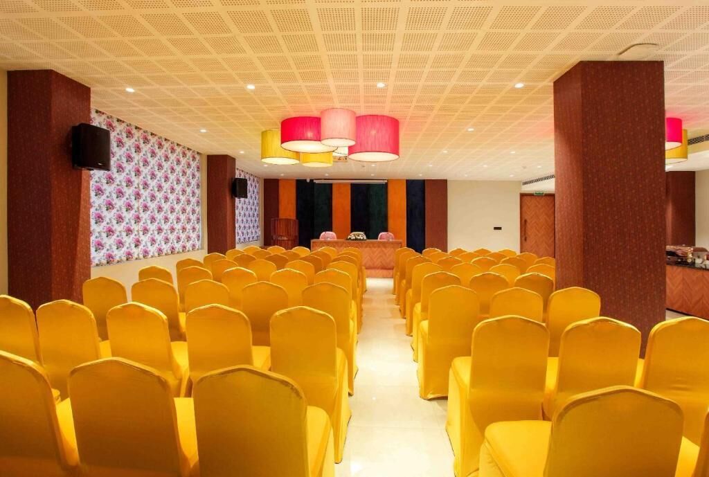 Sala de conferências com fileiras de cadeiras amarelas, palco com um pódio e parede com padrão.