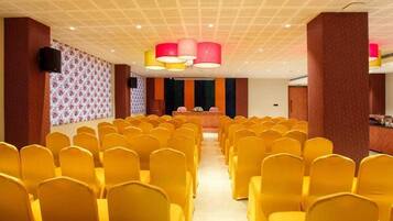 Sala de conferências com fileiras de cadeiras amarelas, palco com um pódio e parede com padrão.