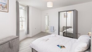 Quarto de hotel moderno com uma cama grande, um banco cinza, um guarda-roupa e uma janela com cortinas.