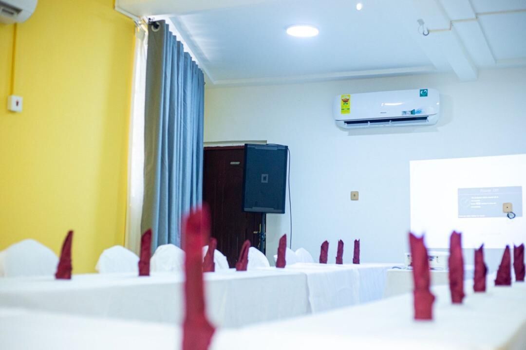 Sala de conferência com mesas brancas, fitas vermelhas, um aparelho de ar condicionado e uma porta fechada.