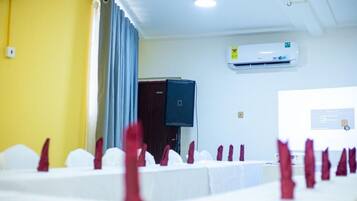 Sala de conferência com mesas brancas, fitas vermelhas, um aparelho de ar condicionado e uma porta fechada.