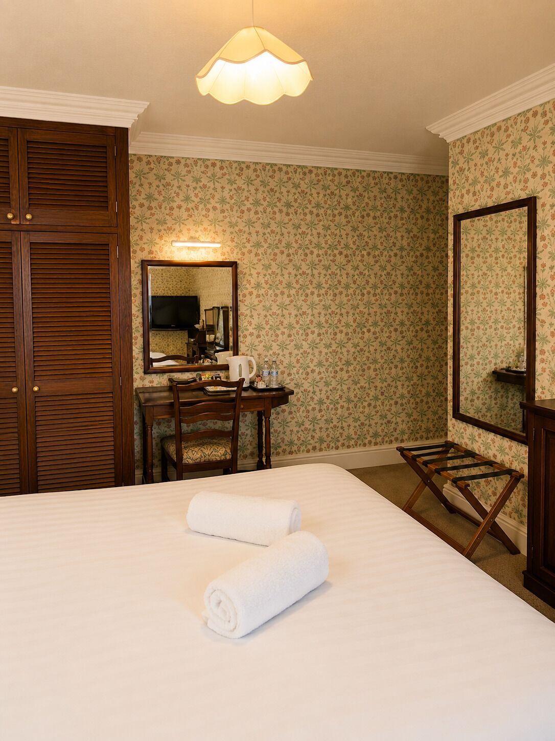 Quarto de hotel com cama, escrivaninha com cadeira, espelho, luminária pendente e guarda-roupa de madeira.