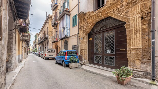 Uma rua estreita ladeada por edifícios históricos, carros estacionados e vasos de plantas.