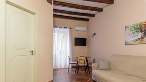 Uma sala de estar com sofá, mesa de jantar com cadeiras, TV fixada na parede e porta que dá para outro cômodo.