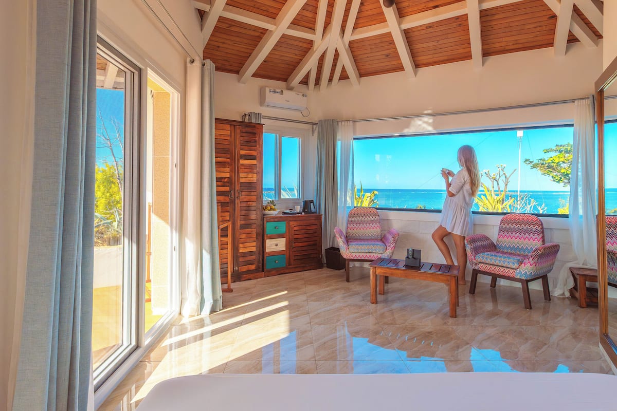Quarto espaçoso com uma grande porta de vidro deslizante que dá para uma varanda com vista para o mar. Há duas cadeiras coloridas e uma mesa pequena.