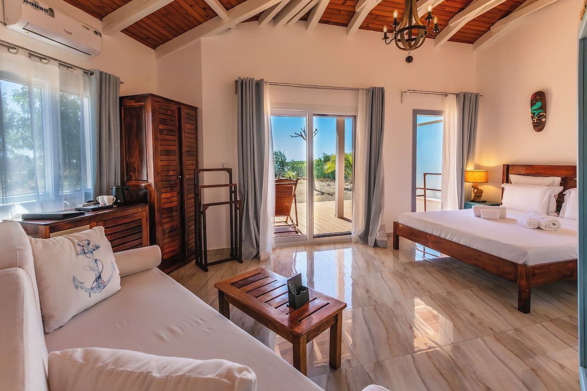 Quarto amplo com cama, sofá, mesa de madeira e escrivaninha. Possui porta de vidro deslizante que dá para área externa com vista para vegetação.