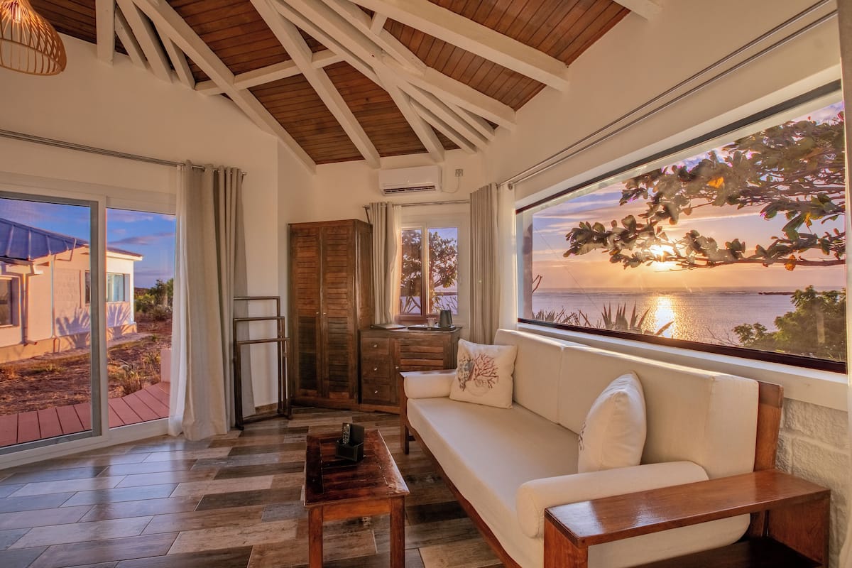 Uma sala de estar acolhedora com teto de madeira, um sofá com almofadas, uma mesa de centro e uma grande janela com vista para um pôr do sol sobre a água.