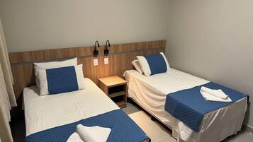 Quarto de hotel com duas camas, cada uma com colcha acolchoada azul e almofadas brancas. Há mesas de cabeceira com luminárias e uma cabeceira de madeira.