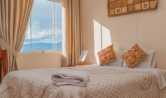 Quarto de hotel com uma cama, janela com cortinas, uma obra de arte na parede e uma cadeira.