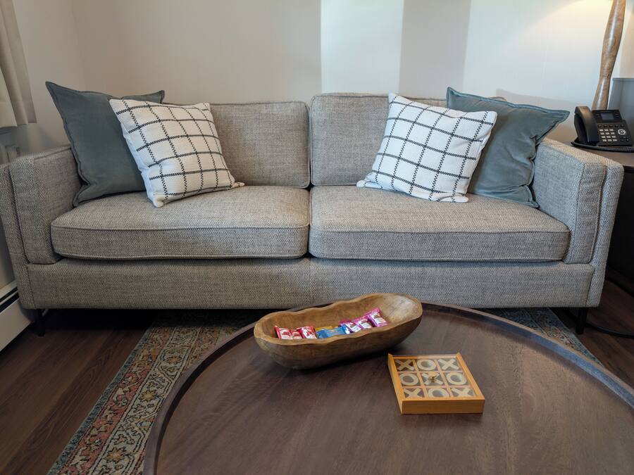 Um sofá cinzento com almofadas estampadas, uma tigela de madeira com petiscos e uma caixa de madeira sobre uma mesa redonda.