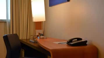 Quarto de hotel com uma escrivaninha, luminária, telefone e cadeira.