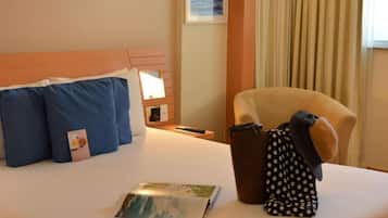 Quarto de hotel com cama, almofadas, uma cadeira, abajur e um livro.