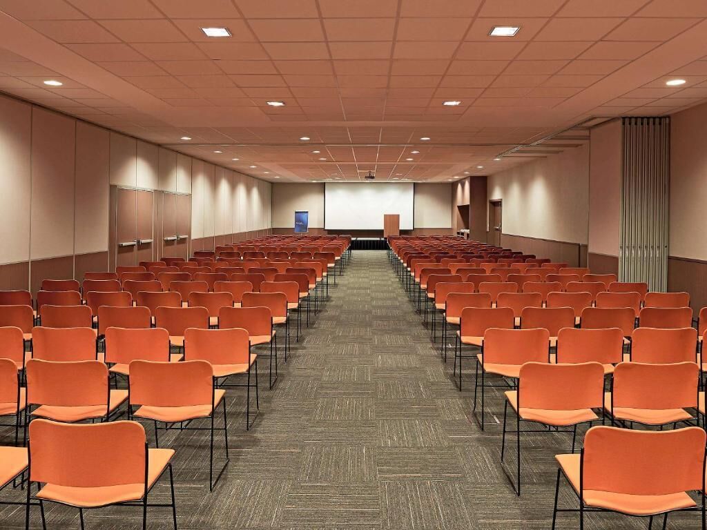 Uma sala de conferências com fileiras de cadeiras laranja, uma tela de projetor e um palanque na frente.