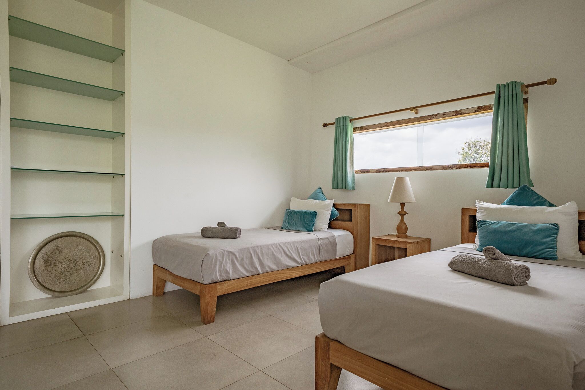 Quarto com duas camas, janela com cortinas, mesa de cabeceira com abajur e estante com um prato.