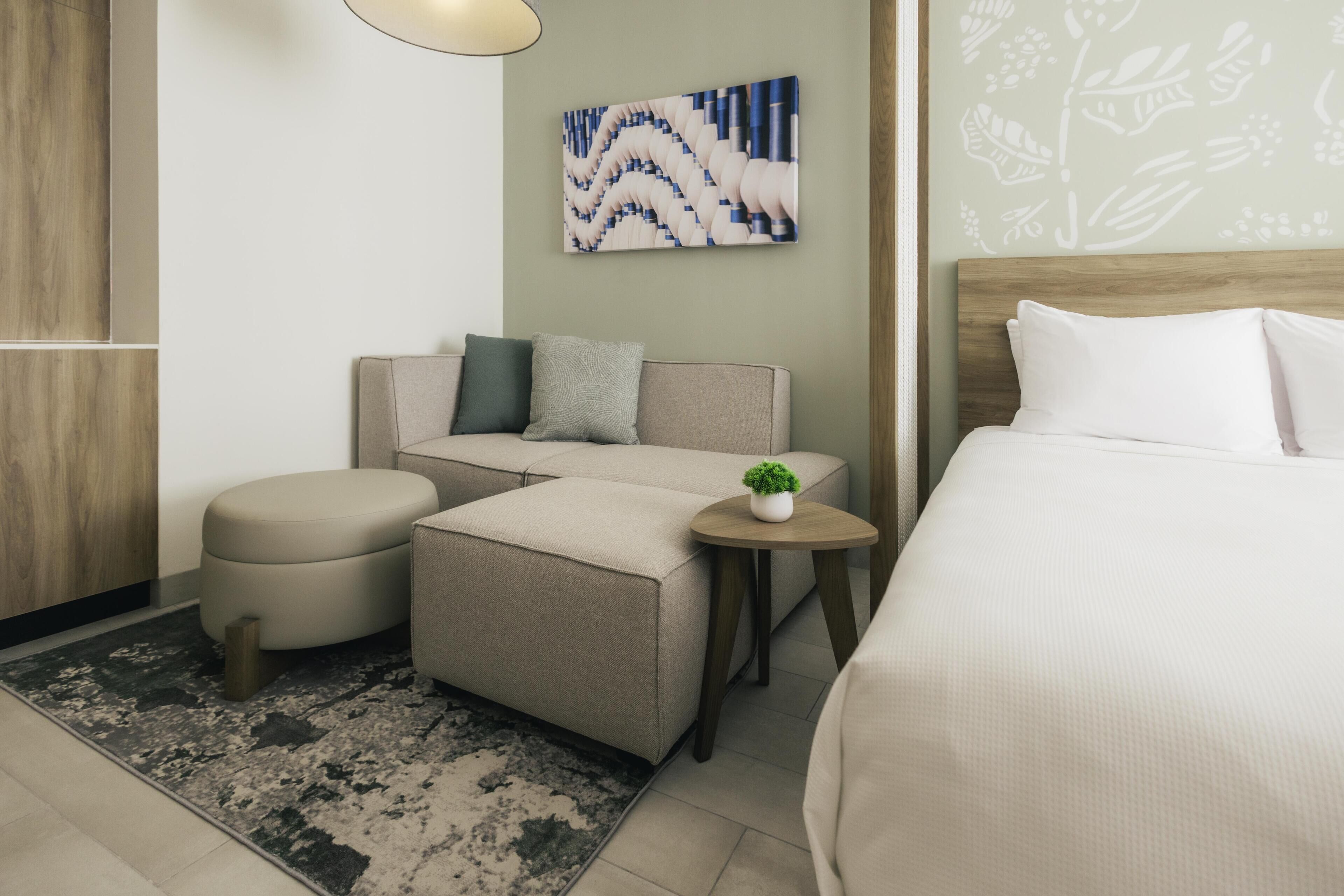 Quarto de hotel moderno com uma cama, um sofá, uma mesinha com uma planta e um objeto decorativo na parede.