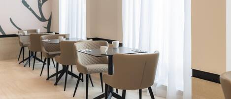 Uma área de refeições moderna com uma mesa comprida e cadeiras, uma parede com design de árvore e grandes janelas com cortinas leves.