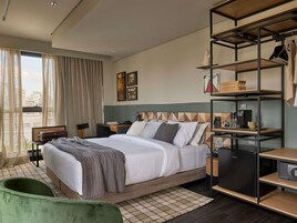 Quarto de hotel moderno com uma cama grande, uma mesa com cafeteira, uma estante de livros e vista para a cidade pela janela.