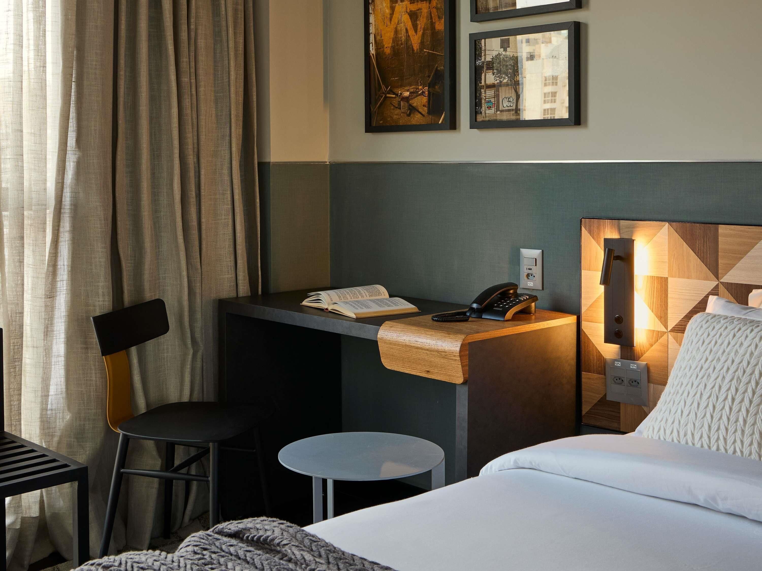 Quarto de hotel com cama, escrivaninha com telefone, cadeira, mesa redonda pequena e luminária fixada na parede.
