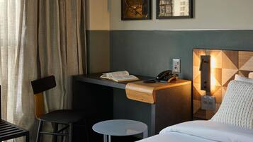 Quarto de hotel com cama, escrivaninha com telefone, cadeira, mesa redonda pequena e luminária fixada na parede.