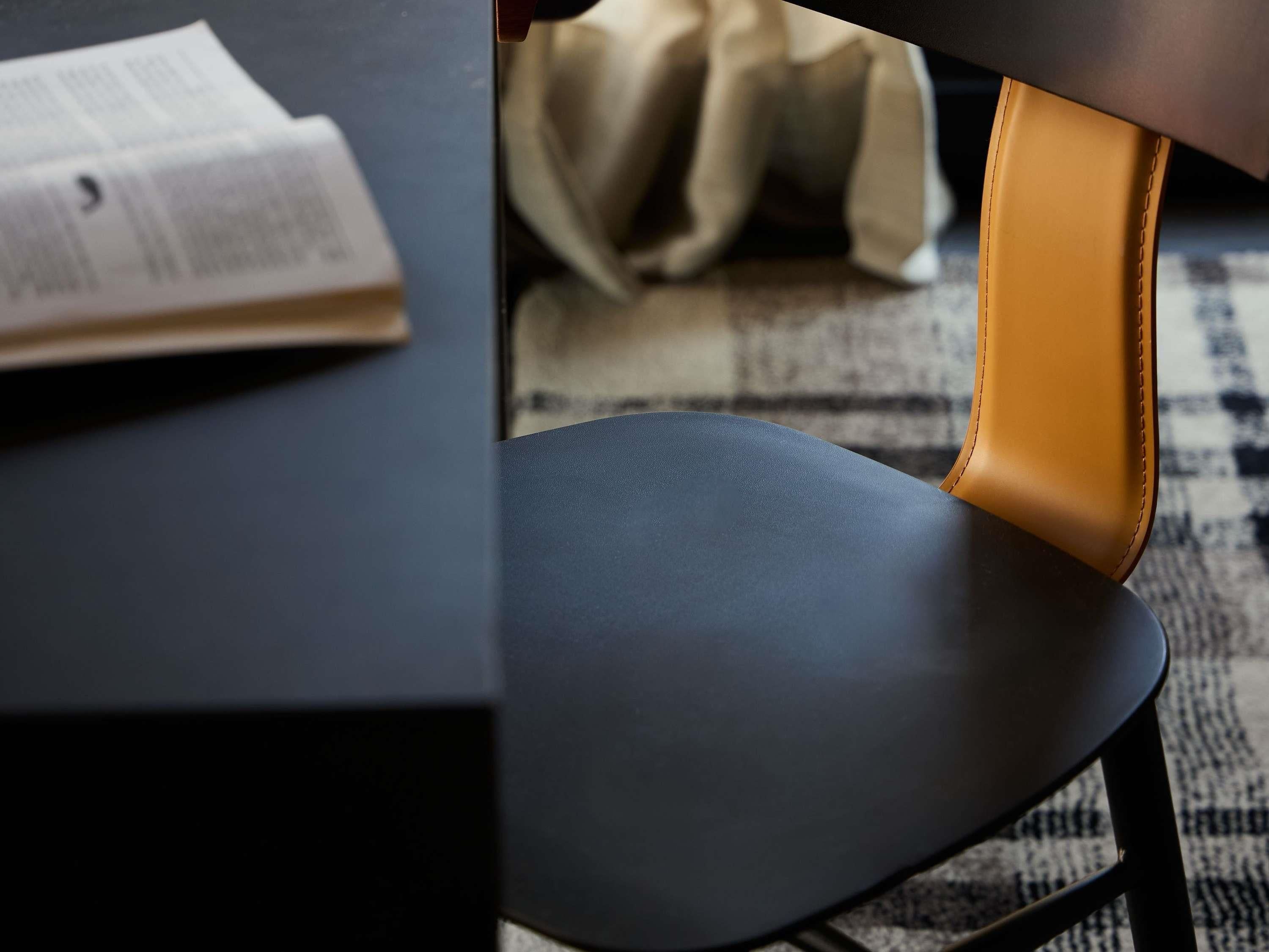 Uma cadeira com assento escuro e encosto cor de bronze, posicionada em frente a uma mesa com um livro.