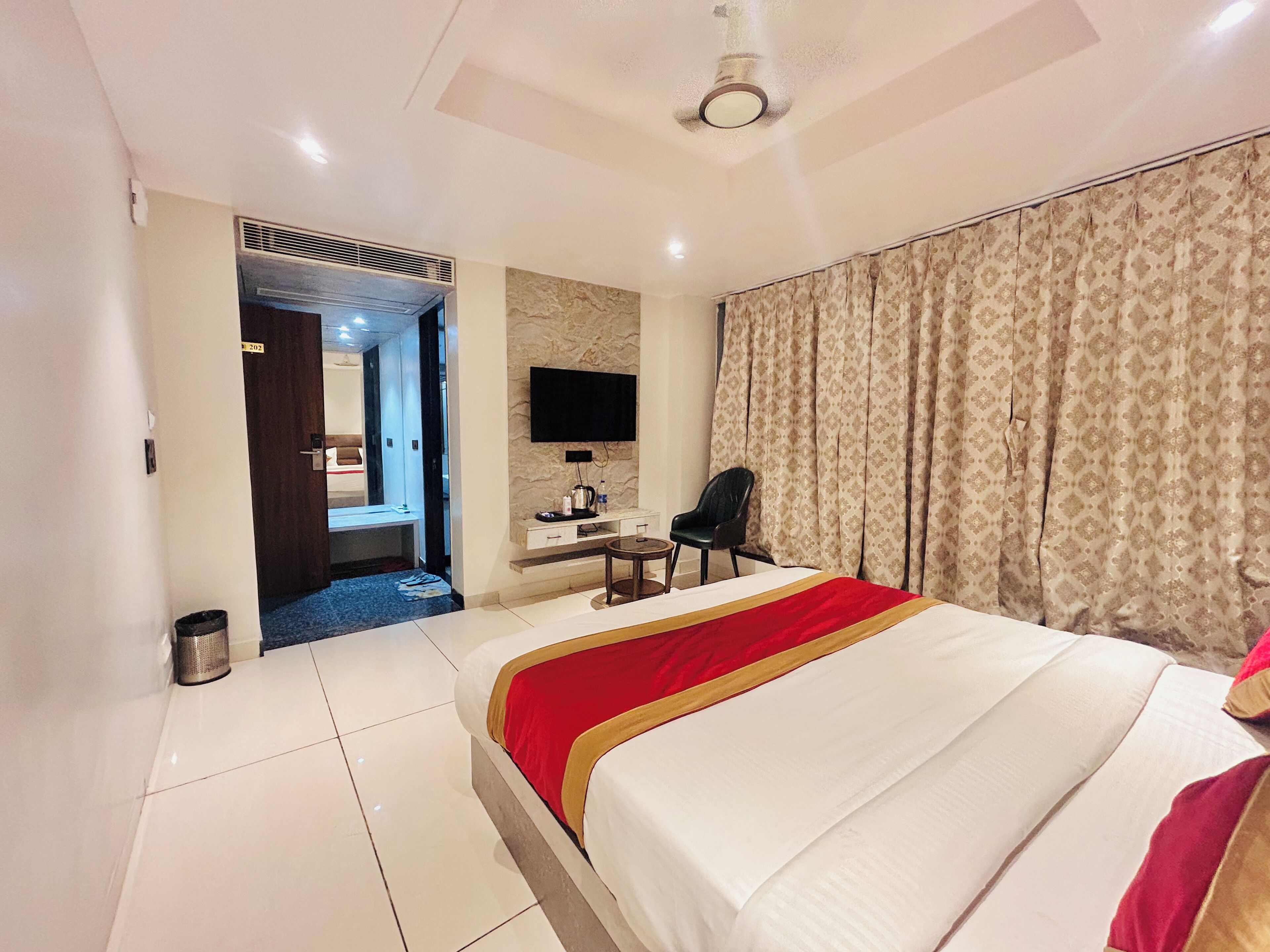 Quarto de hotel com uma cama grande, uma escrivaninha com cadeira, uma televisão fixada na parede e um banheiro visível através de uma porta aberta.