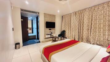 Quarto de hotel com uma cama grande, uma escrivaninha com cadeira, uma televisão fixada na parede e um banheiro visível através de uma porta aberta.