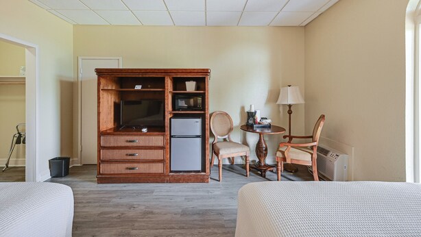 Quarto de hotel com guarda-roupa de madeira, frigorífico, uma cadeira, mesa com abajur e uma cama.