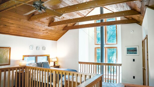 Quarto com teto inclinado, vigas de madeira, cama com almofadas, mesa de cabeceira com abajur e janela com vista para árvores.