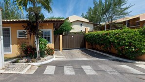 Edifício residencial com portão fechado, entrada por um caminho pavimentado e um pequeno jardim com plantas e flores.