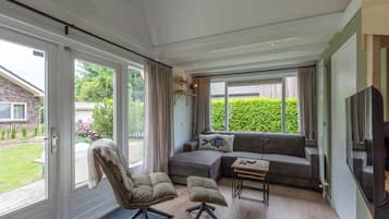 Uma sala de estar moderna com um sofá seccional, uma mesinha e uma cadeira. Há uma televisão na parede e uma porta de vidro deslizante que dá para um jardim.