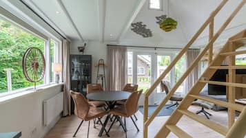 Uma área de refeições com uma mesa redonda e cadeiras, um radiador, uma janela com vista para vegetação e uma escadaria de madeira.