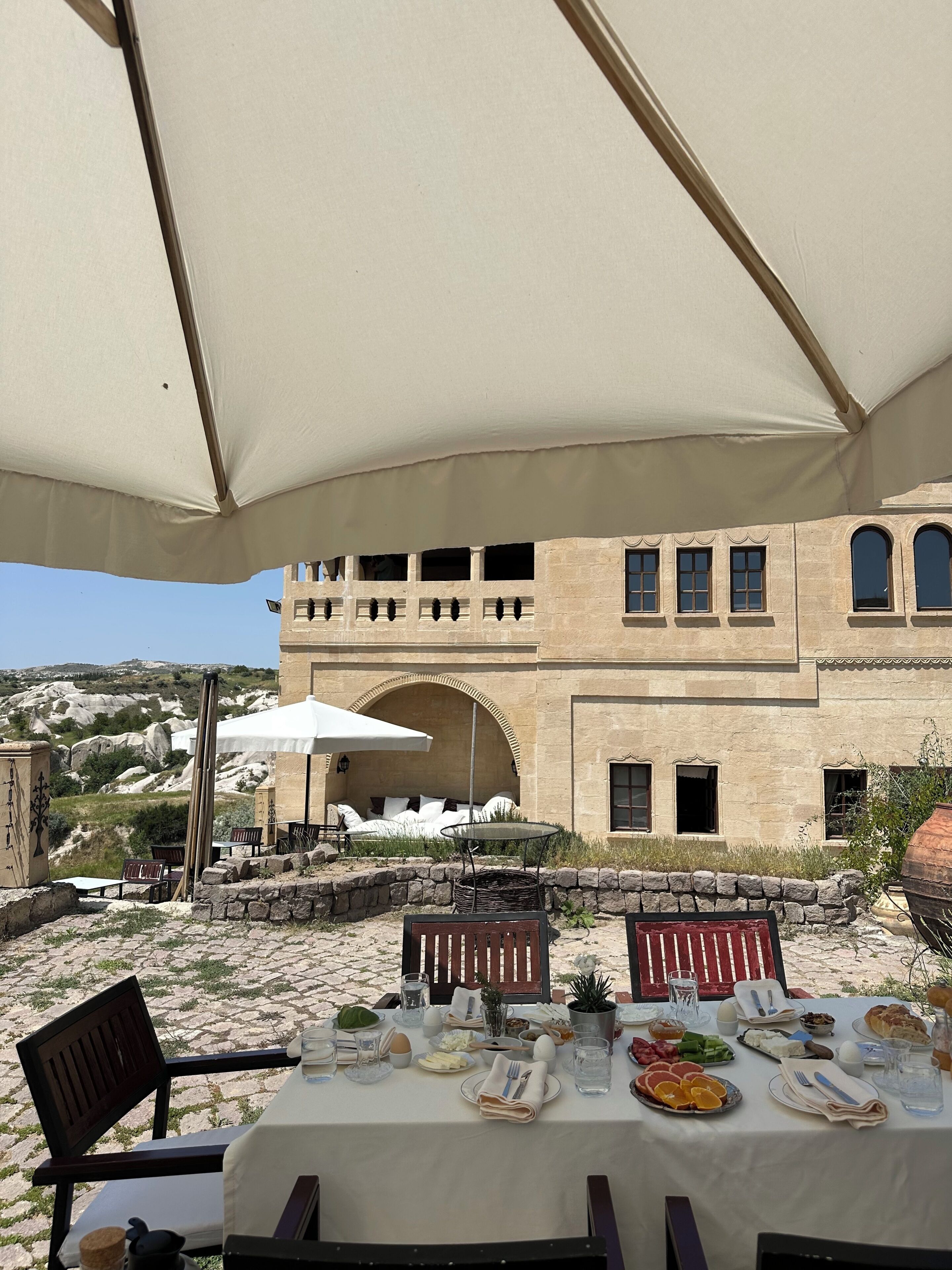 Uma área de refeições ao ar livre com uma mesa posta, um guarda-sol e um edifício com janelas em arco ao fundo.