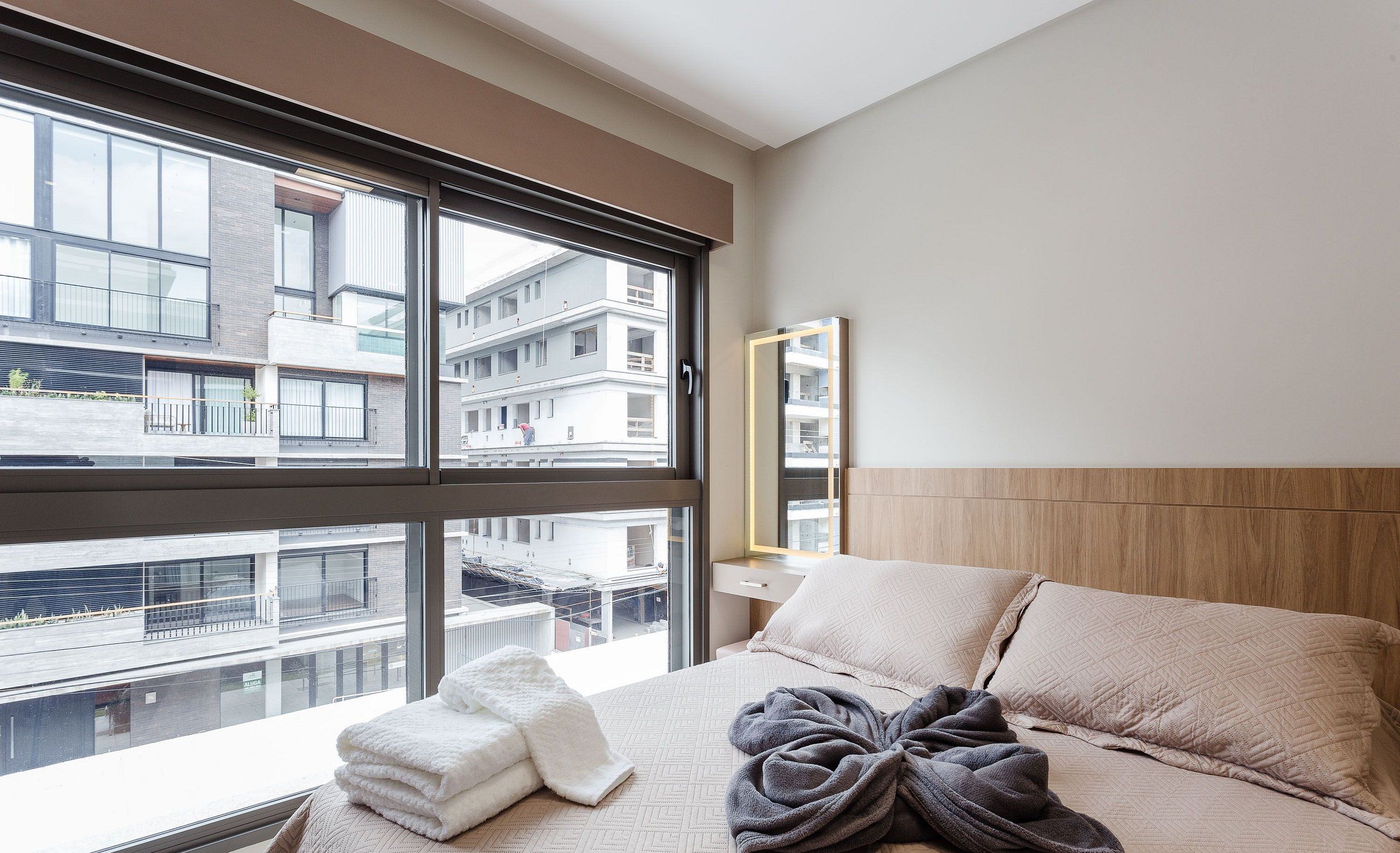 Quarto de hotel moderno com uma janela grande, cama com colcha bege, cabeceira de madeira e vista para outros edifícios.