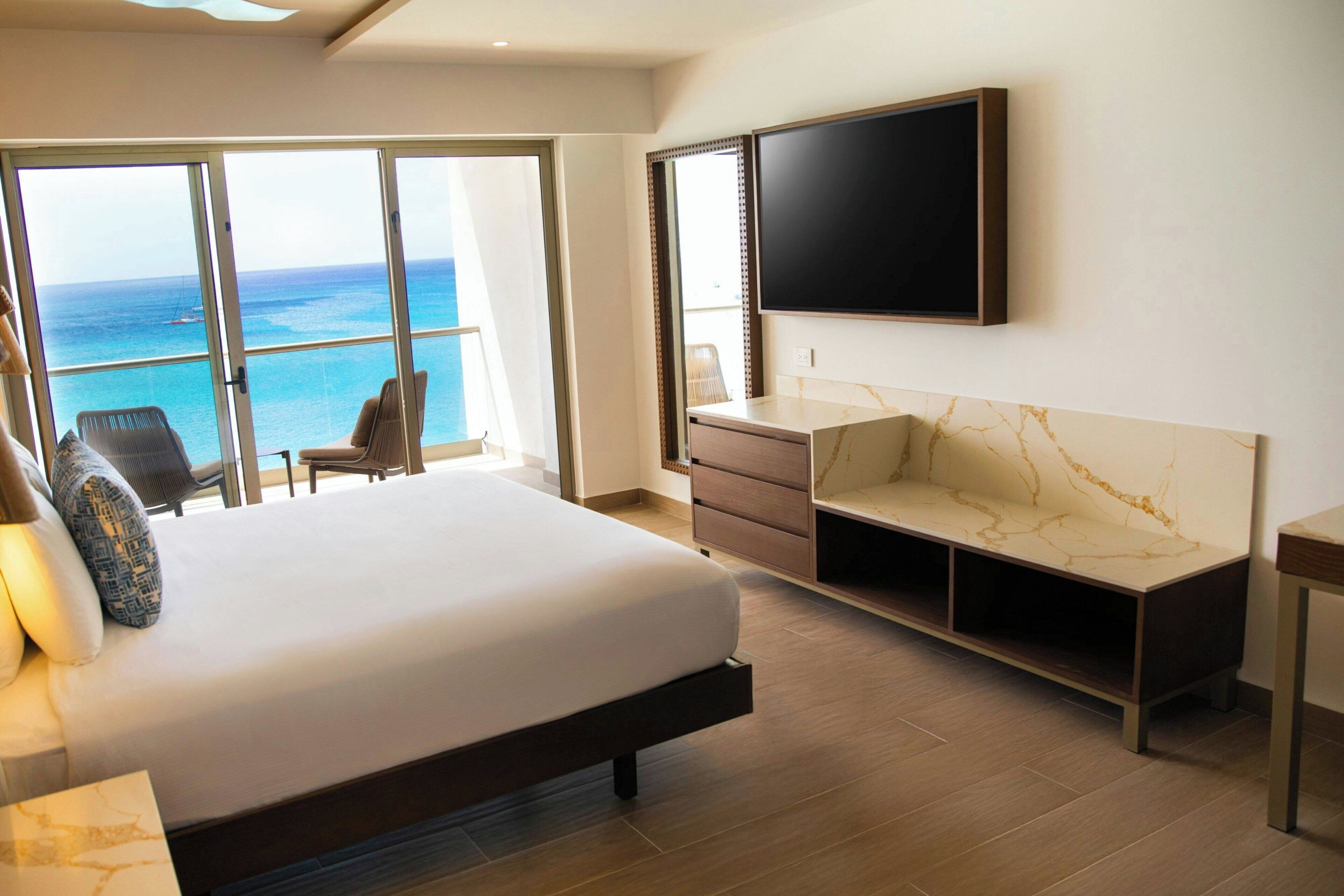 Quarto com cama grande, televisão na parede, escrivaninha e vista para o oceano.