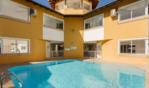 Uma área de piscina com piscina azul cristalina, corrimãos brancos e fachada de prédio amarela.