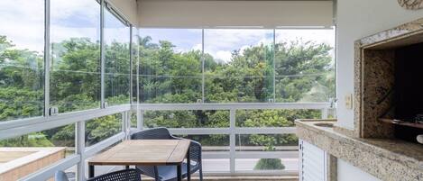 Uma varanda com mesa e cadeiras de madeira, área de cozinha com bancada e janelas amplas com vista para vegetação.
