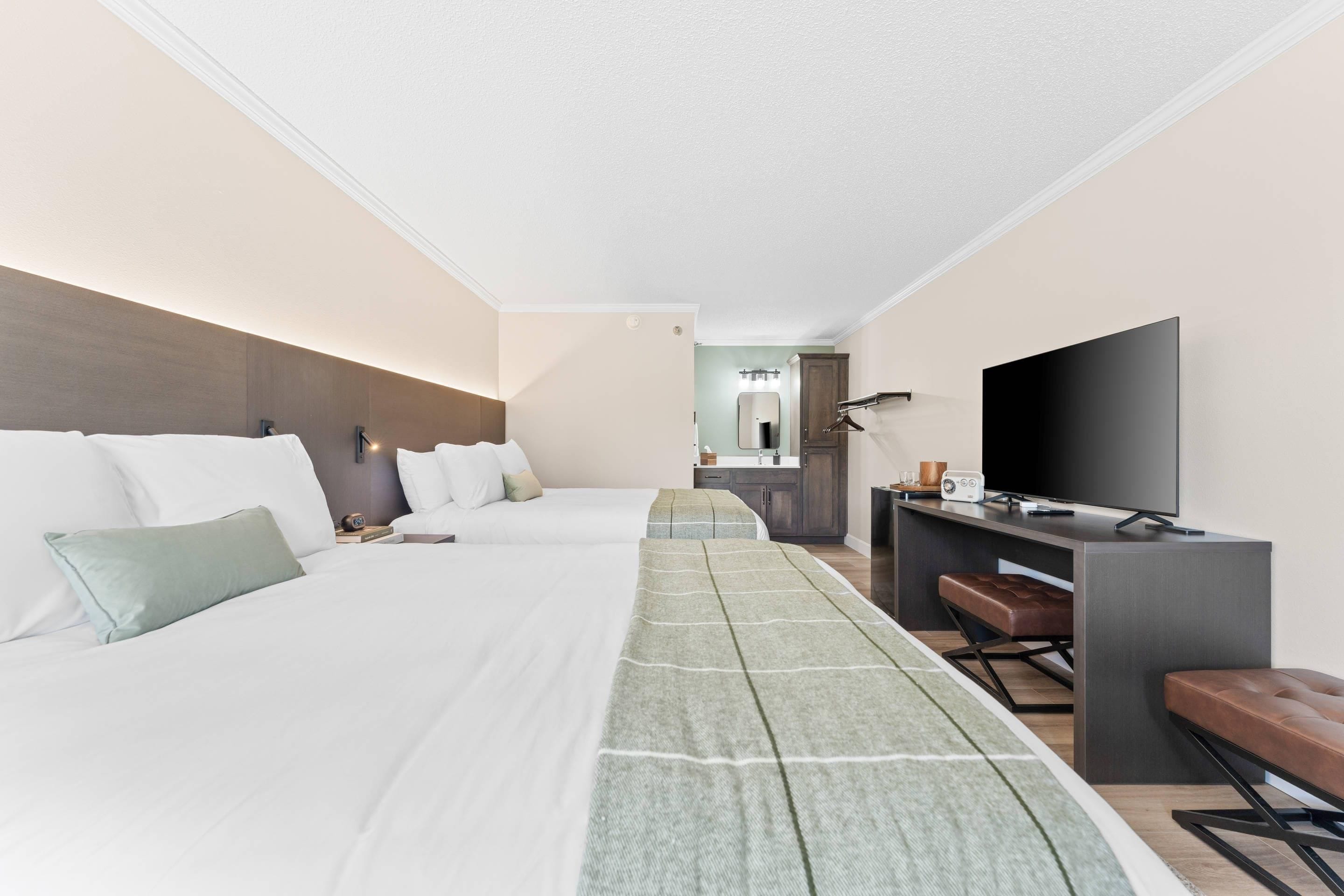 Quarto de hotel com cama, escrivaninha com televisão, um banco e banheiro ao fundo.