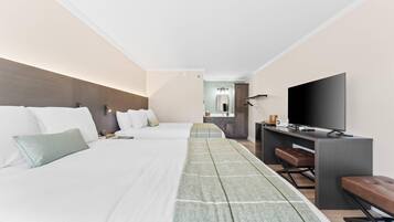 Quarto de hotel com cama, escrivaninha com televisão, um banco e banheiro ao fundo.