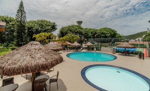 Área de piscina com duas piscinas, guarda-sóis de palha, espreguiçadeiras e vista para um tanque d'água.