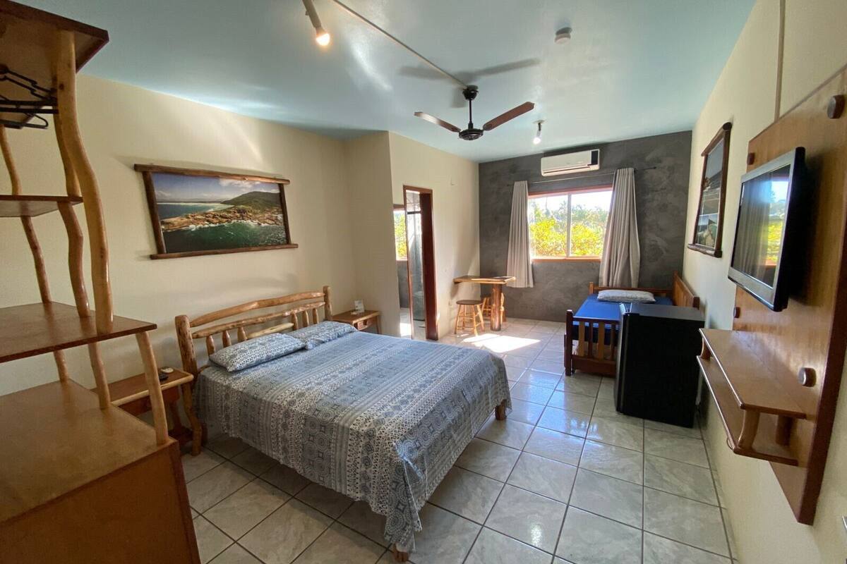 Quarto com cama, escrivaninha, cadeira, ventoinha no teto e televisão.