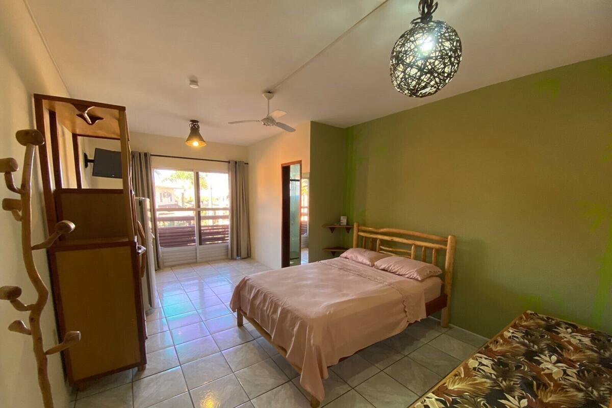Quarto com cama, ventilador de teto, luminária pendente e vista para área externa por uma porta de correr.