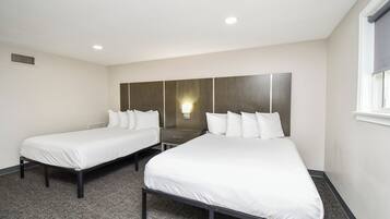 Quarto de hotel com duas camas de solteiro, uma mesa de cabeceira, uma luminária na parede e uma janela com cortina.