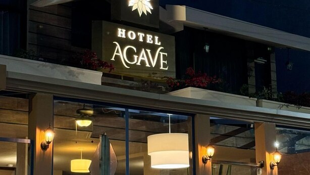 Um hotel chamado "Hotel Agave" com um letreiro iluminado chamativo, janelas amplas e vista para as montanhas ao fundo.