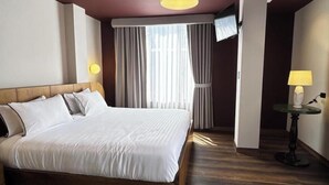 Quarto de hotel com uma cama grande, piso de madeira, televisão fixada na parede e luminária sobre uma mesinha.