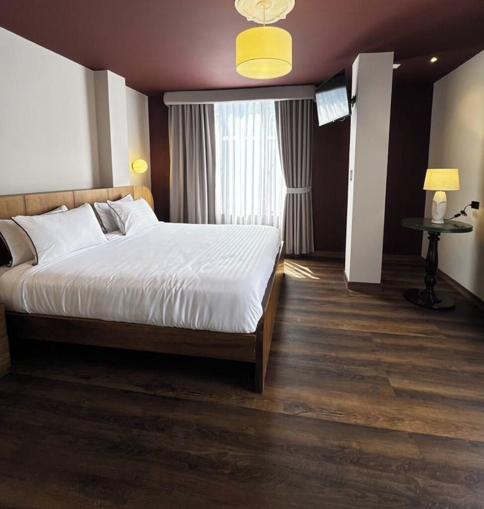 Quarto de hotel com uma cama grande, piso de madeira, televisão fixada na parede e luminária sobre uma mesinha.
