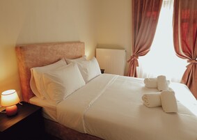 Cama bem arrumada com roupa de cama e almofadas brancas, uma luminária de cabeceira e uma janela com cortinas.