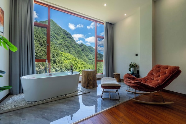 Casa de banho moderna com uma banheira independente, uma cadeira de balanço vermelha e uma janela ampla com vista para uma paisagem montanhosa.