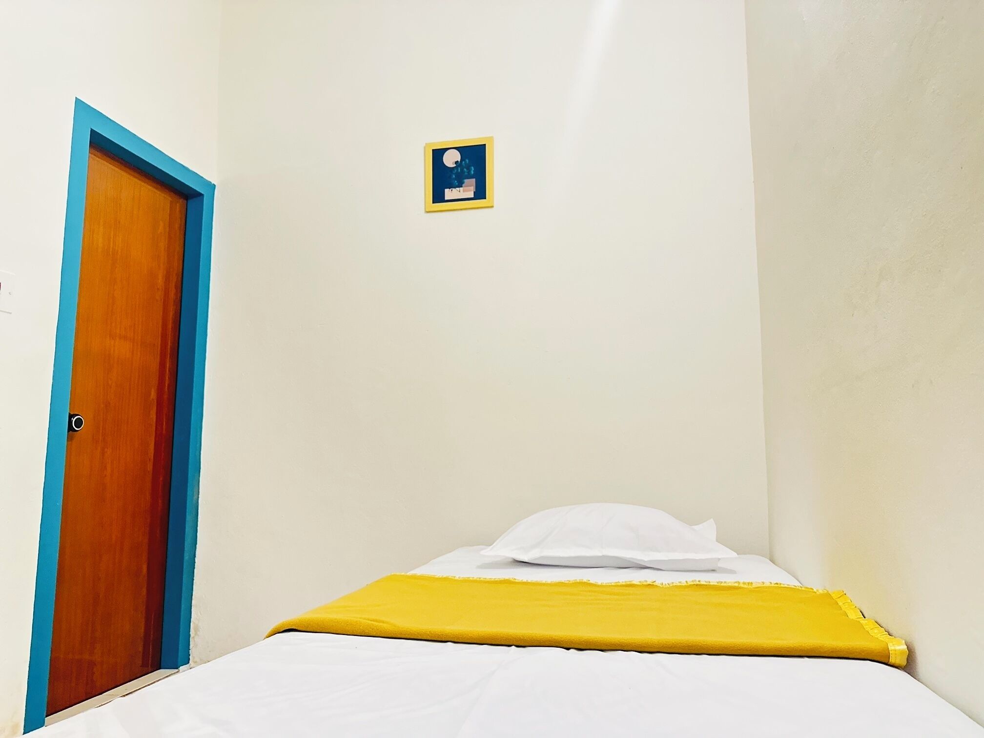 Quarto pequeno com uma cama de solteiro, porta de madeira com moldura azul e um pequeno quadro na parede.