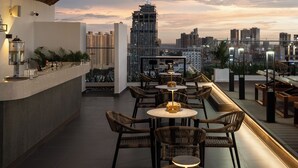 Um bar no terraço com mobiliário de vime, balcão de bar e vista para a cidade ao pôr do sol.